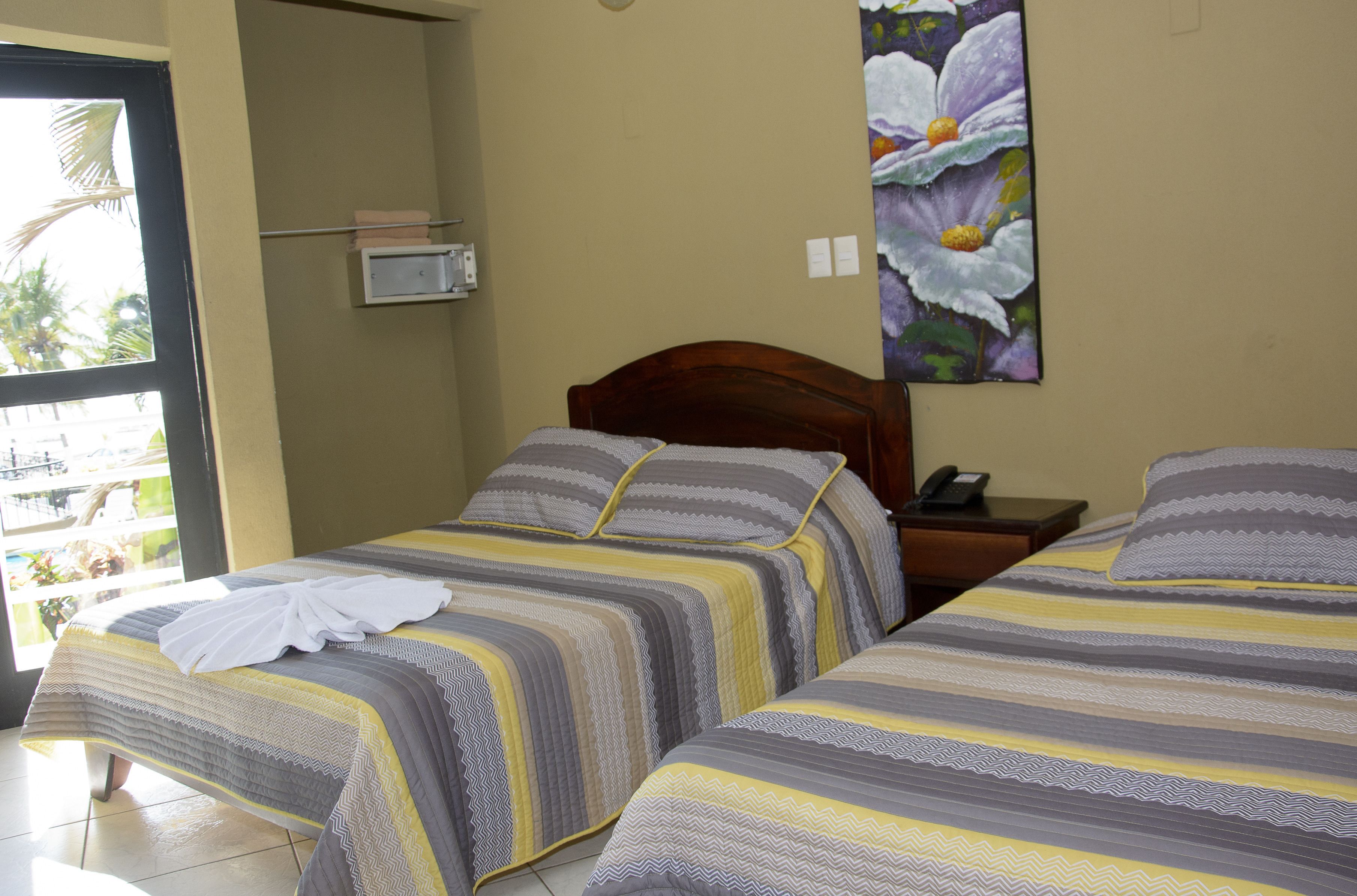 Quarto de hotel com duas camas, cabeceira de madeira, mesa de cabeceira, quadro de flores e janela com vista para palmeiras.