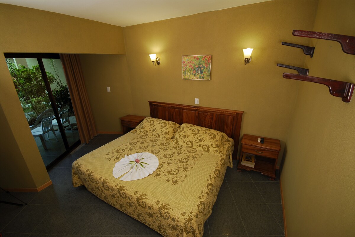 Quarto de hotel com cama, duas mesas de cabeceira, uma mesinha de apoio, porta de correr de vidro para uma varanda e duas luminárias fixadas na parede.