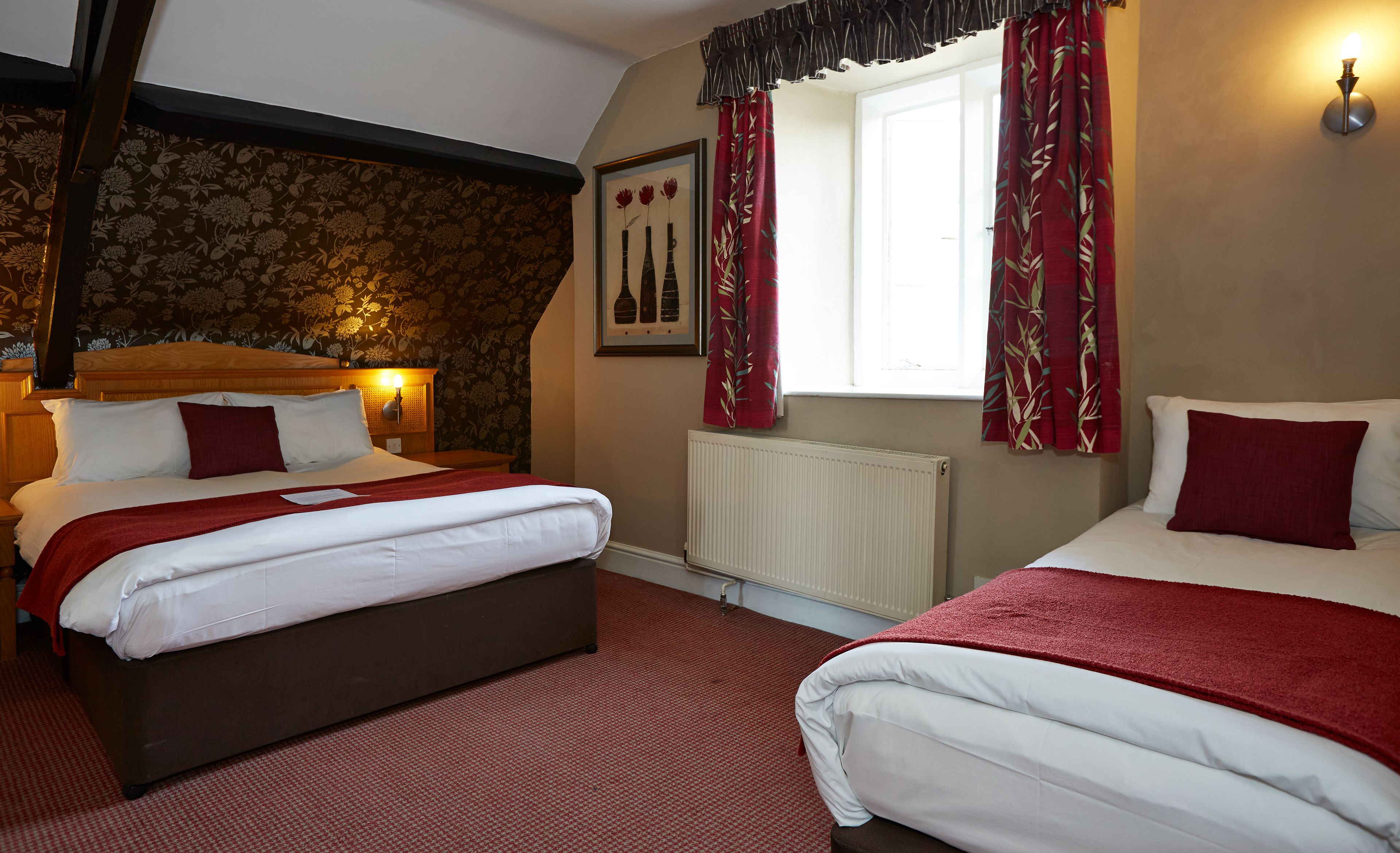 Quarto de hotel com duas camas, um radiador, uma janela com cortinas, parede com papel de parede floral e um quadro emoldurado sobre a cama.