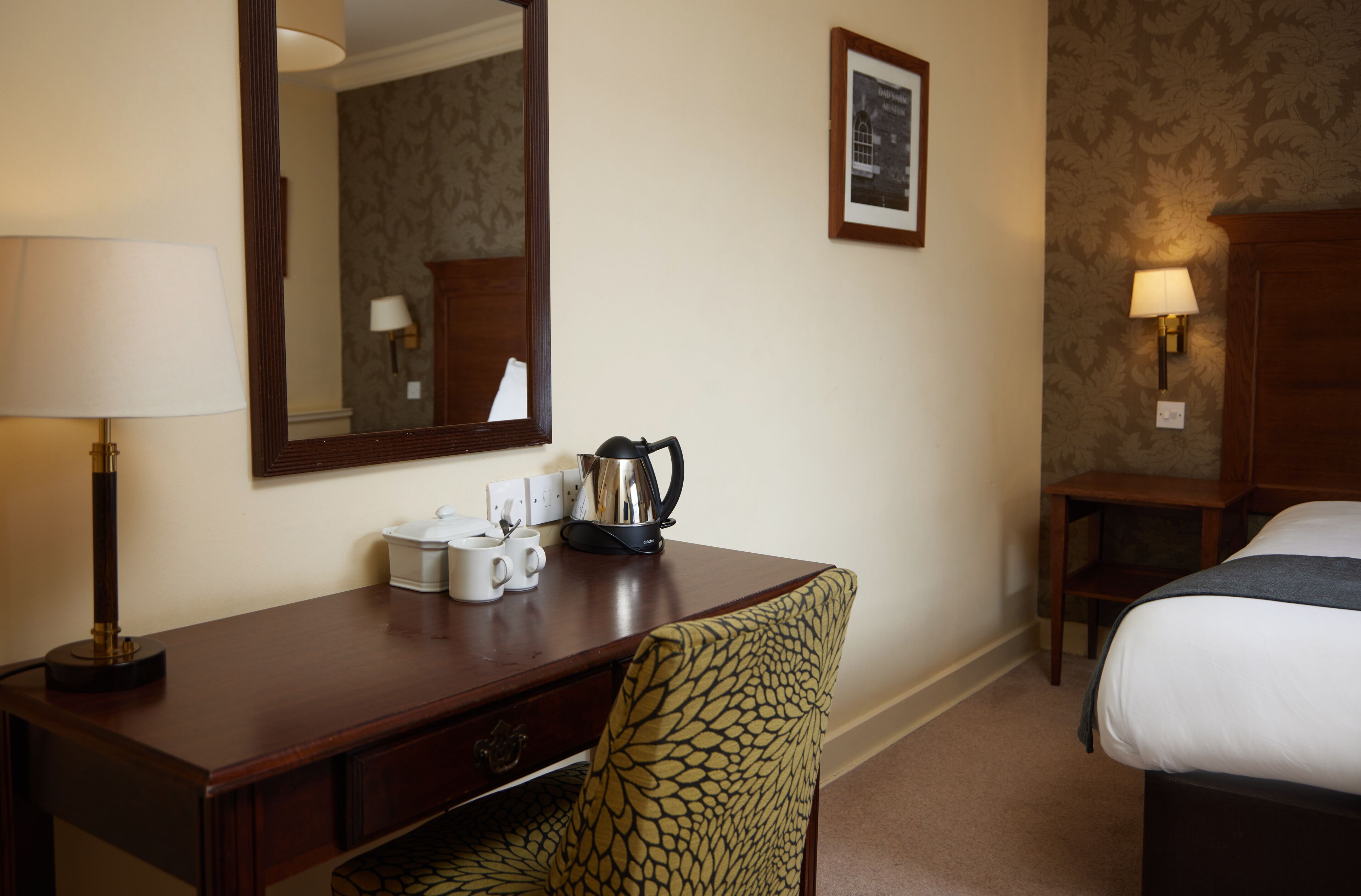 Quarto de hotel com escrivaninha de madeira, uma cadeira, cama e mesa de cabeceira.