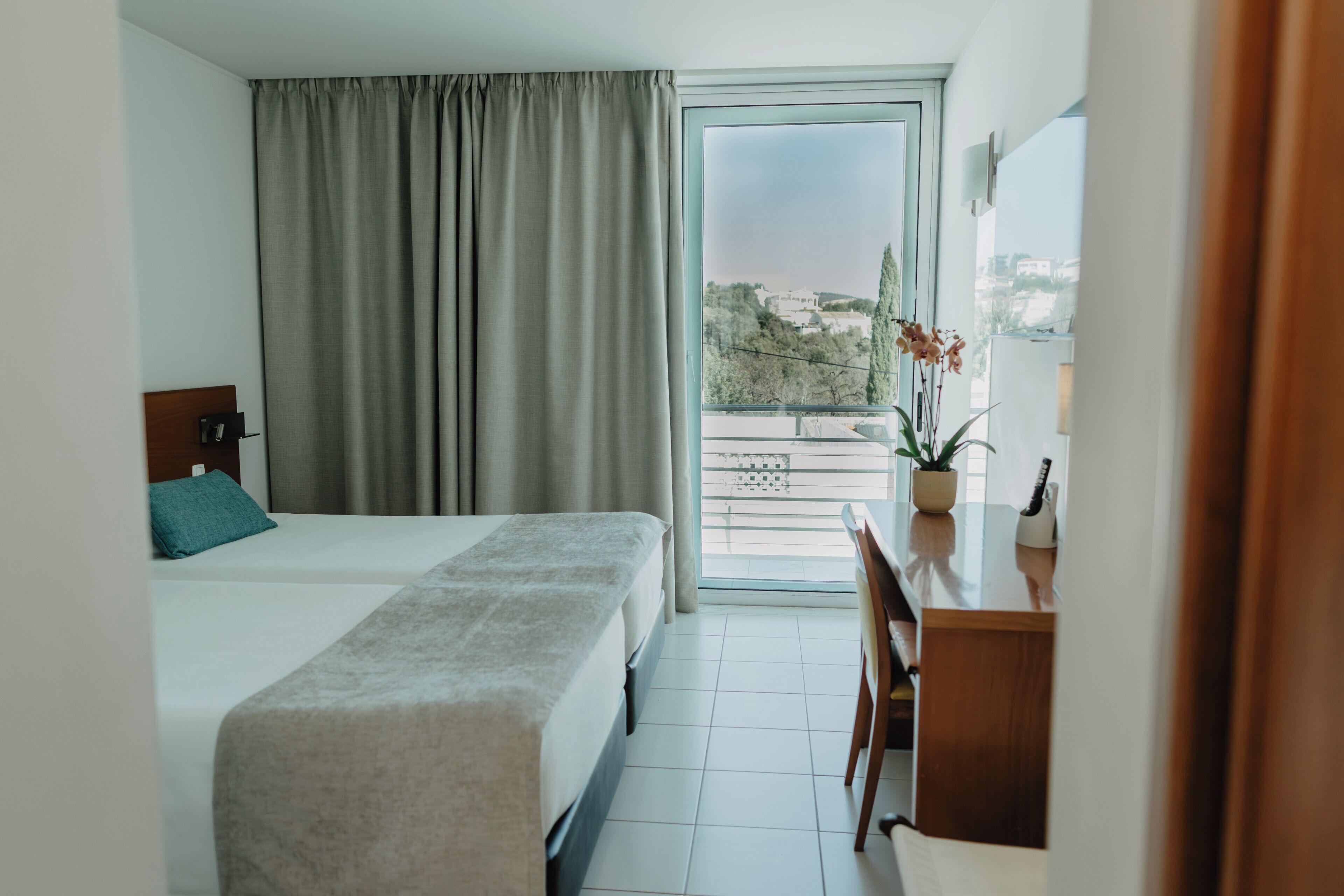 Quarto de hotel com cama, escrivaninha, cadeira, planta em vaso e varanda com vista.