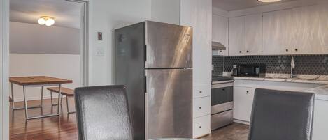 Cozinha moderna com eletrodomésticos de aço inoxidável, sala de jantar com mesa e cadeiras de vidro, e micro-ondas à vista.