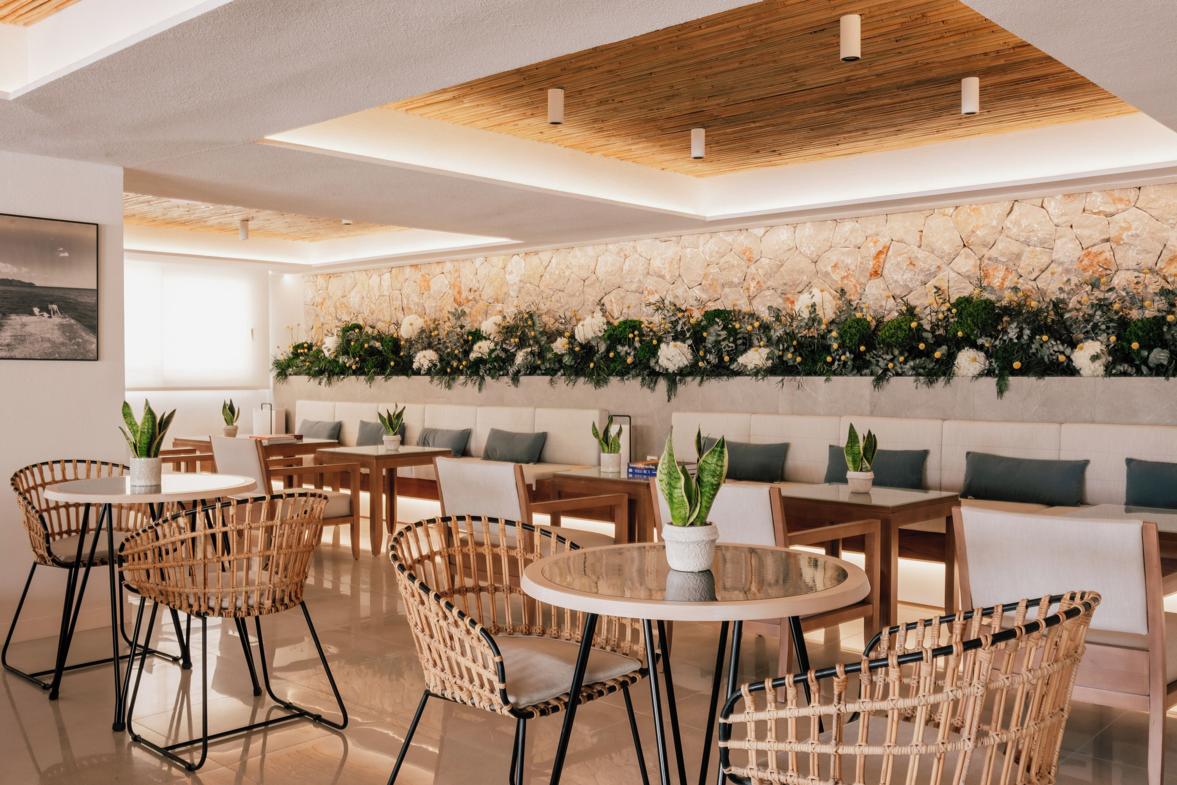 Um interior moderno com mesas redondas, cadeiras de vime e uma parede de pedra decorada com plantas.