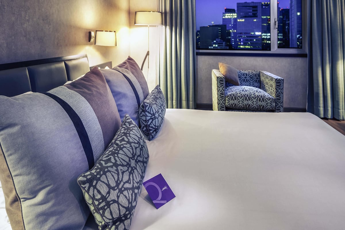Quarto de hotel com cama, uma cadeira, vista para a cidade e janela com cortinas.