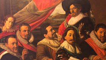 Uma pintura histórica retrata um grupo de pessoas reunidas em volta de uma mesa, algumas segurando livros e outras conversando.