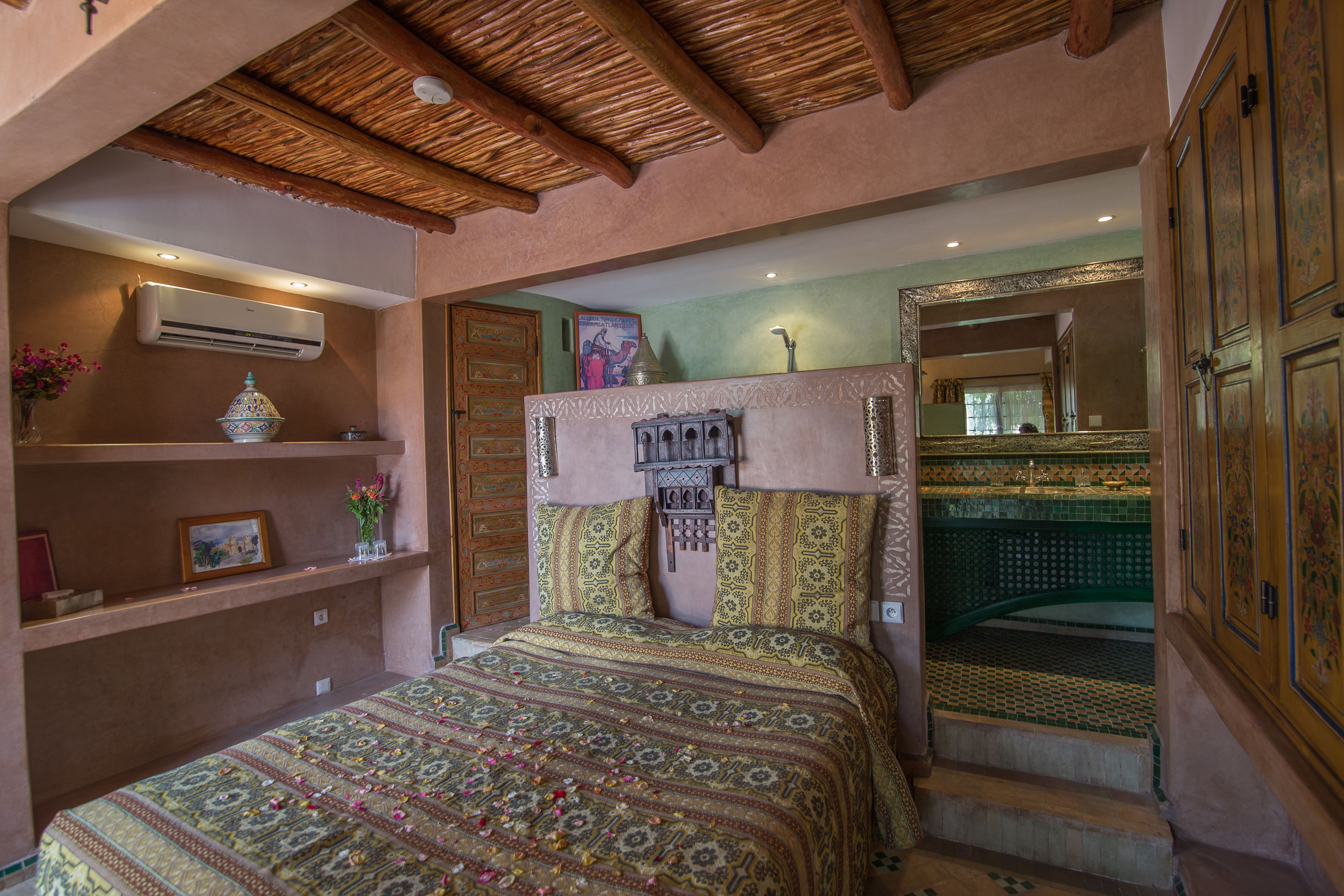 Quarto com colcha estampada, ar condicionado fixado na parede, prateleira com objetos decorativos e um espelho refletindo um banheiro com piso ladrilhado.