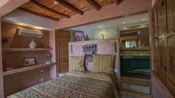 Quarto com colcha estampada, ar condicionado fixado na parede, prateleira com objetos decorativos e um espelho refletindo um banheiro com piso ladrilhado.