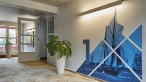 Um corredor com piso de azulejos, uma planta em vaso, uma porta e uma parede com o número '1' grande e uma pintura náutica.