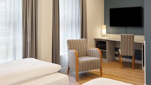 Quarto de hotel com cama, escrivaninha com cadeira, televisão e cortinas.