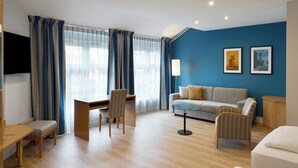 Quarto de hotel moderno com escrivaninha, cadeira, sofá e poltrona, com uma janela grande e cortinas.