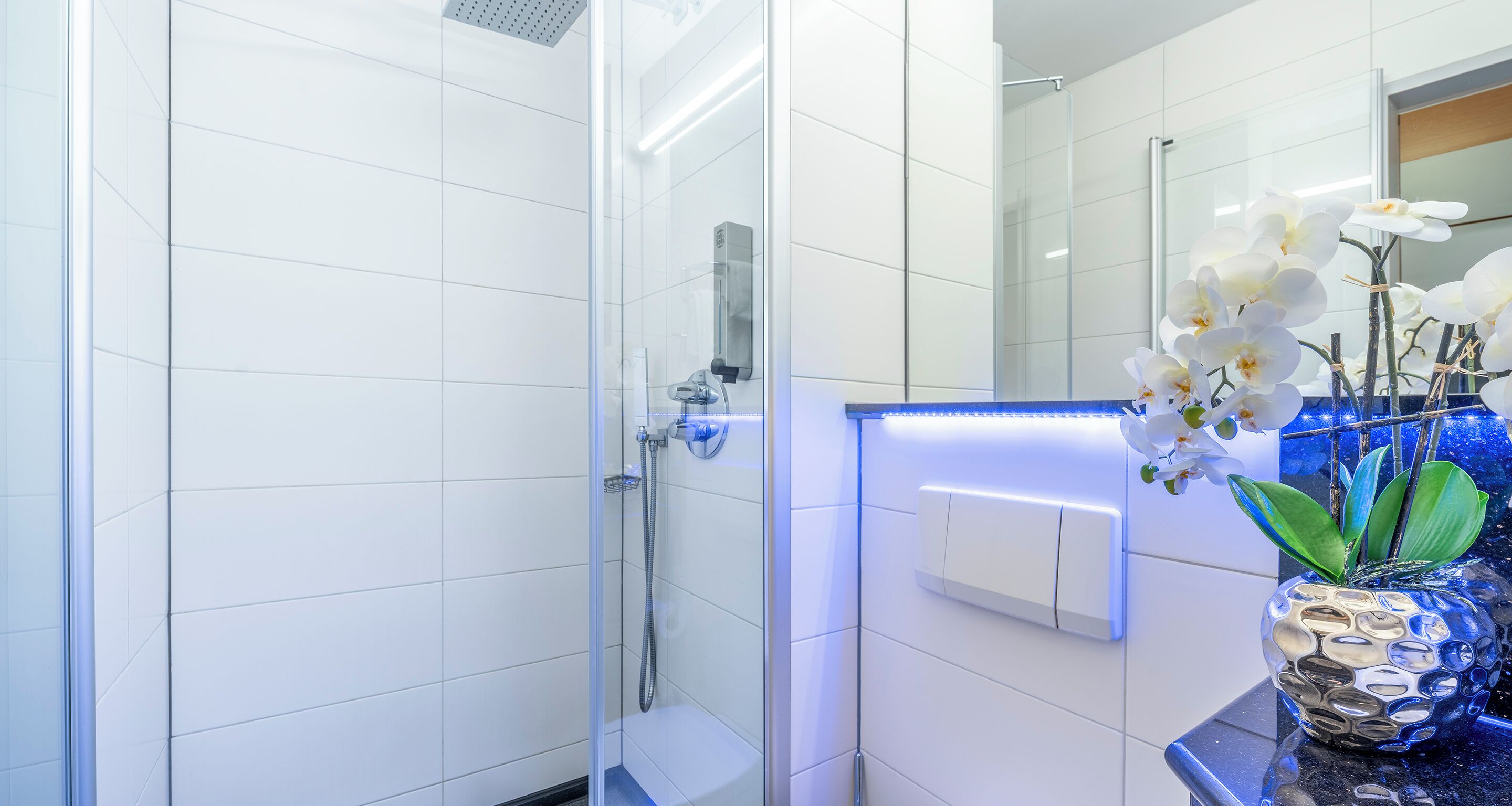 Um banheiro moderno com paredes revestidas de azulejos brancos, box de vidro, porta-toalhas, vaso sanitário e uma orquídea ornamental em vaso azul.