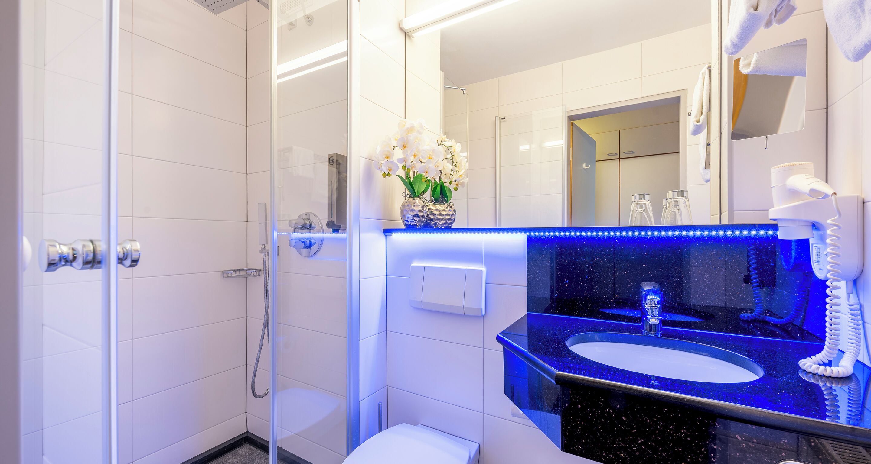 Uma casa de banho moderna com cabine de duche em vidro, lavatório preto com iluminação LED azul e sanita branca.