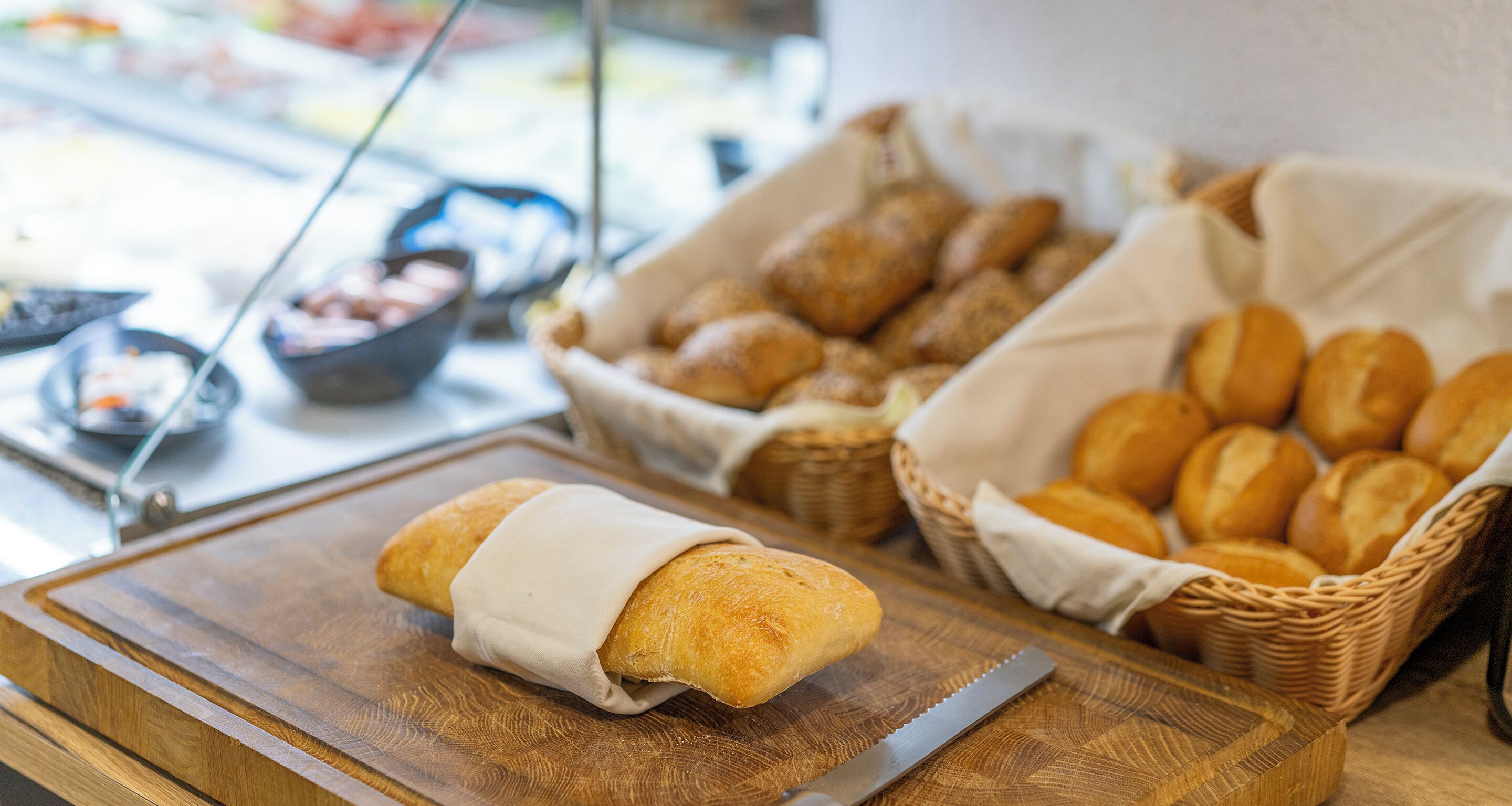 Tábua de madeira com um doce embalado, uma faca e cestos de pão ao fundo.