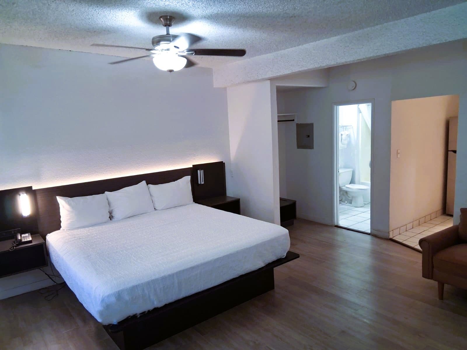 Quarto com uma cama grande, um ventilador de teto, uma mesa de cabeceira e uma porta para um banheiro.