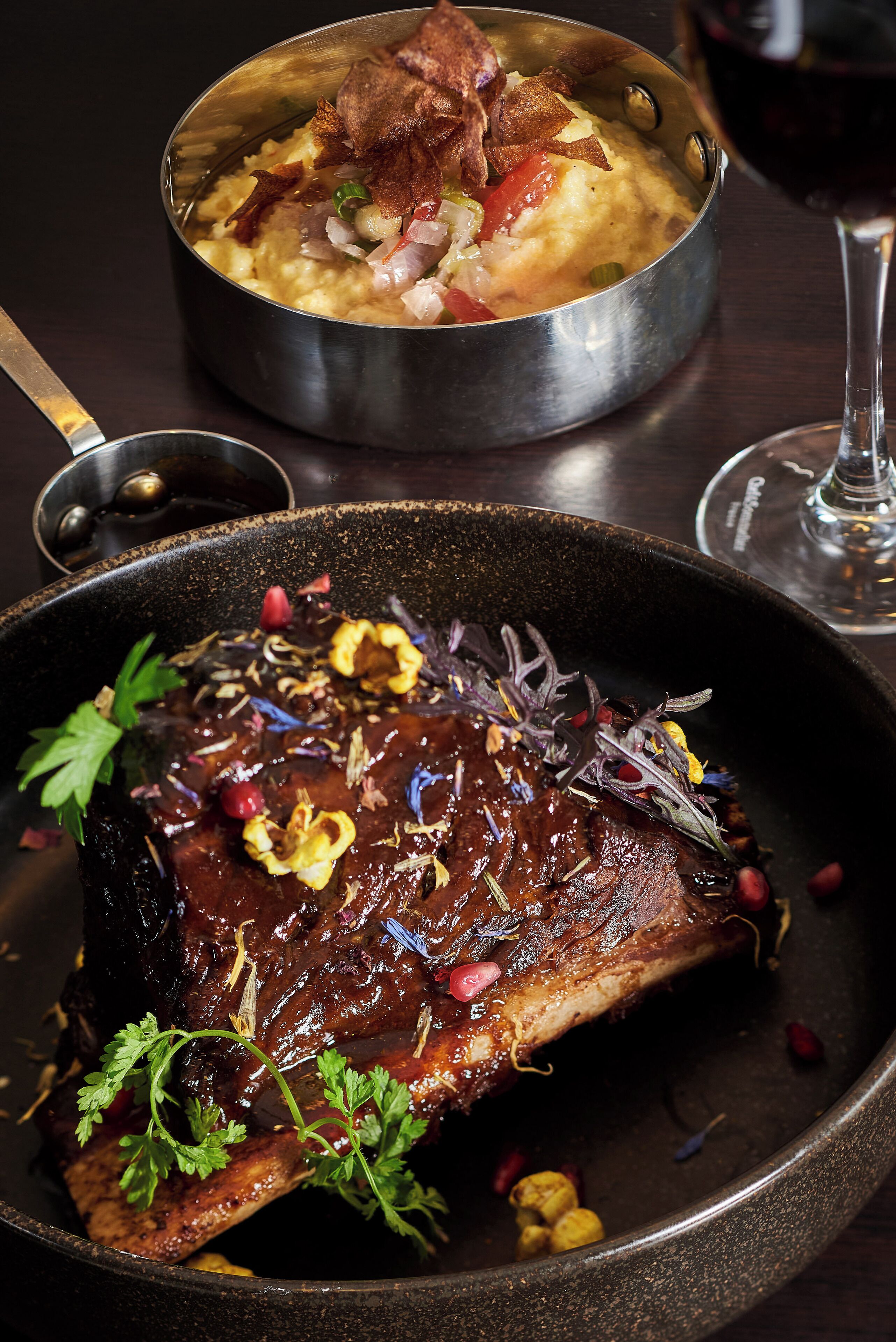 Prato com carne como peça central, coberta por um glace, decorado com ervas e flores comestíveis, acompanhado de um refogado de batatas com vegetais e um copo de vinho tinto.