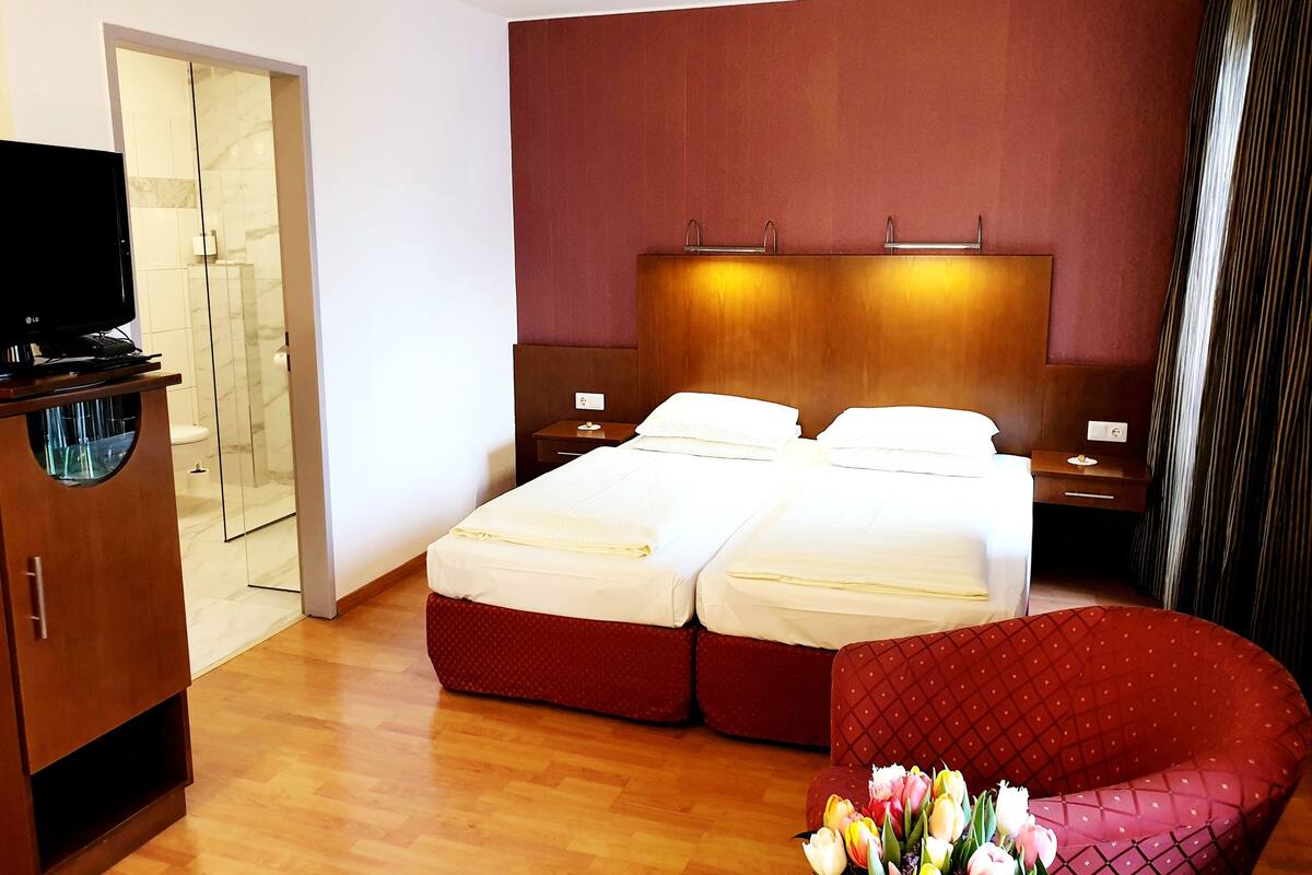Quarto de hotel com cabeceira de madeira, duas camas, uma televisão, um guarda-roupa e uma poltrona vermelha.