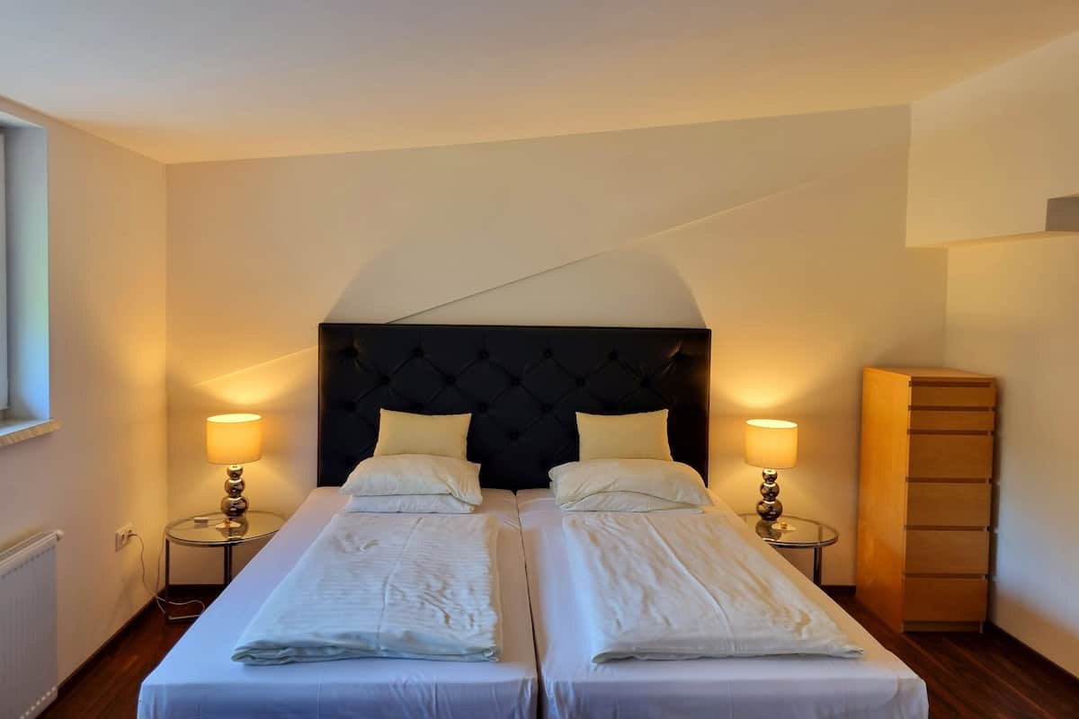 Uma cama de casal com cabeceira escura, duas mesas de cabeceira com luminárias e um armário de madeira.