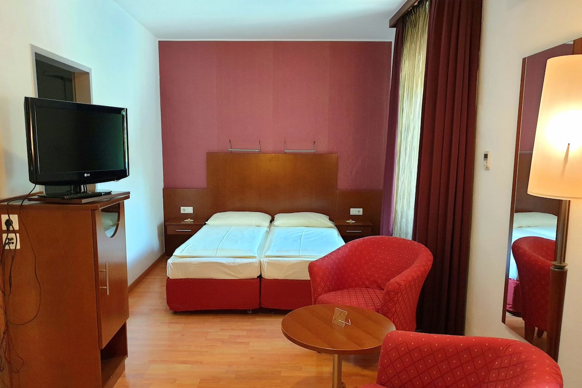 Quarto de hotel com uma cama, uma televisão sobre um móvel, uma mesinha e uma poltrona vermelha.