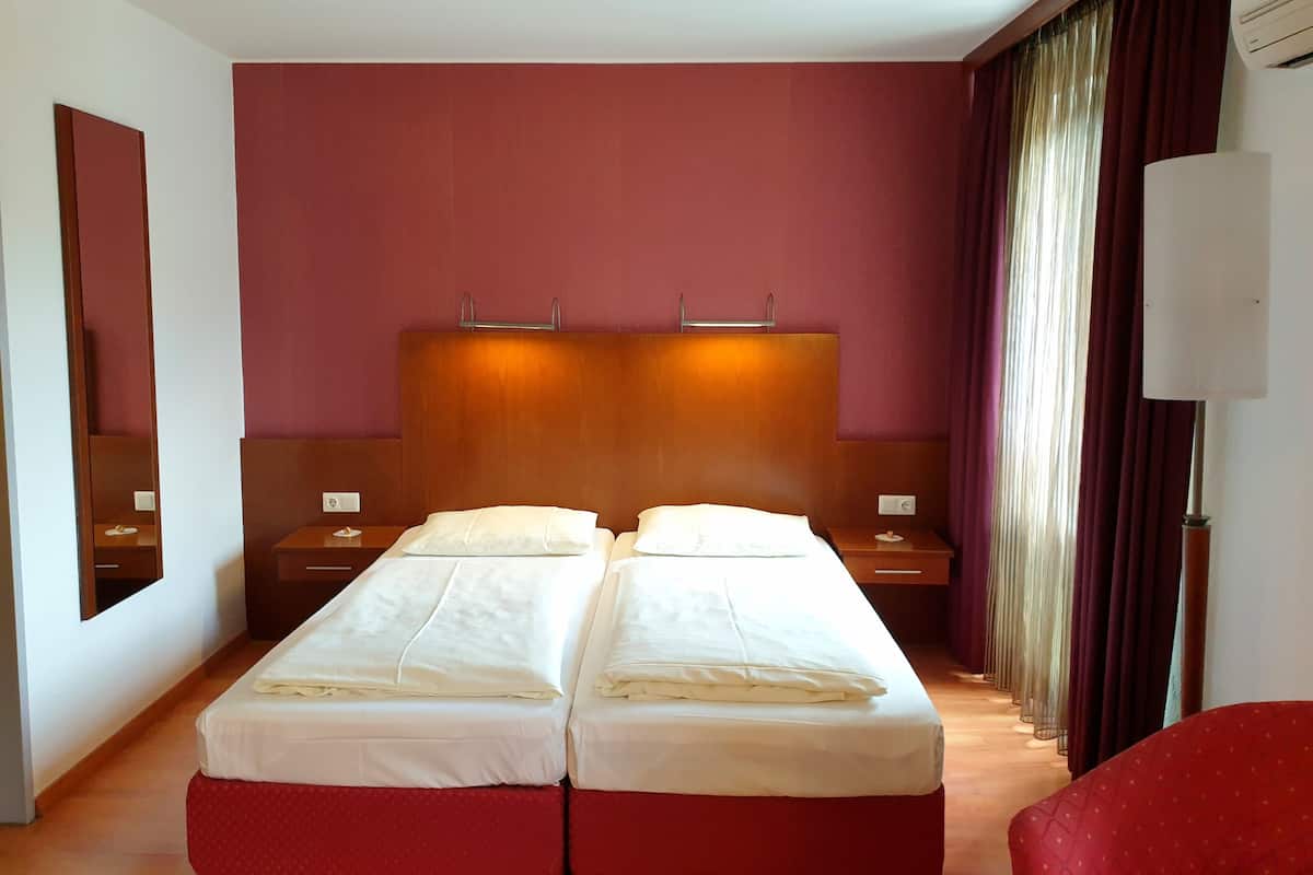 Quarto de hotel com duas camas, cabeceira de madeira, espelho, mesa de cabeceira e uma cadeira vermelha.