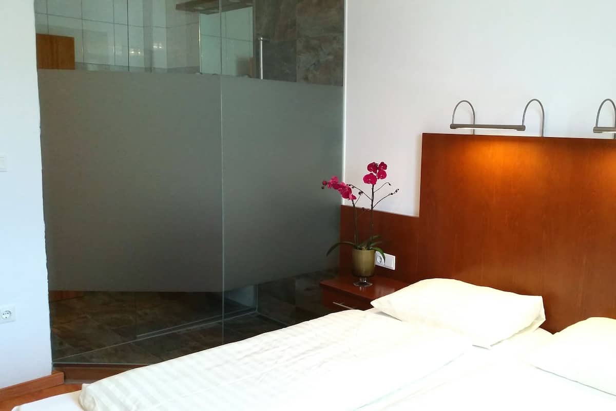 Quarto de hotel moderno com chuveiro em cabine de vidro, painel de cama de madeira e mesa de cabeceira com um vaso de flores cor-de-rosa.
