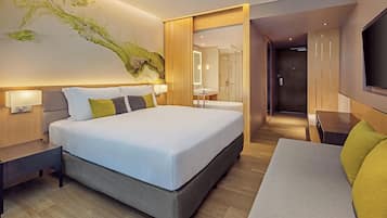 Quarto de hotel moderno com uma cama grande, mesas de cabeceira, um sofá, banheiro com chuveiro e uma TV fixada na parede.