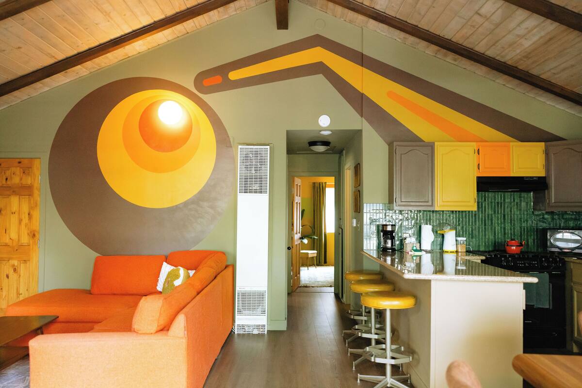 Uma cozinha acolhedora e moderna, com uma ilha central, um espaço de refeições com um sofá laranja vibrante e uma grande decoração circular na parede.