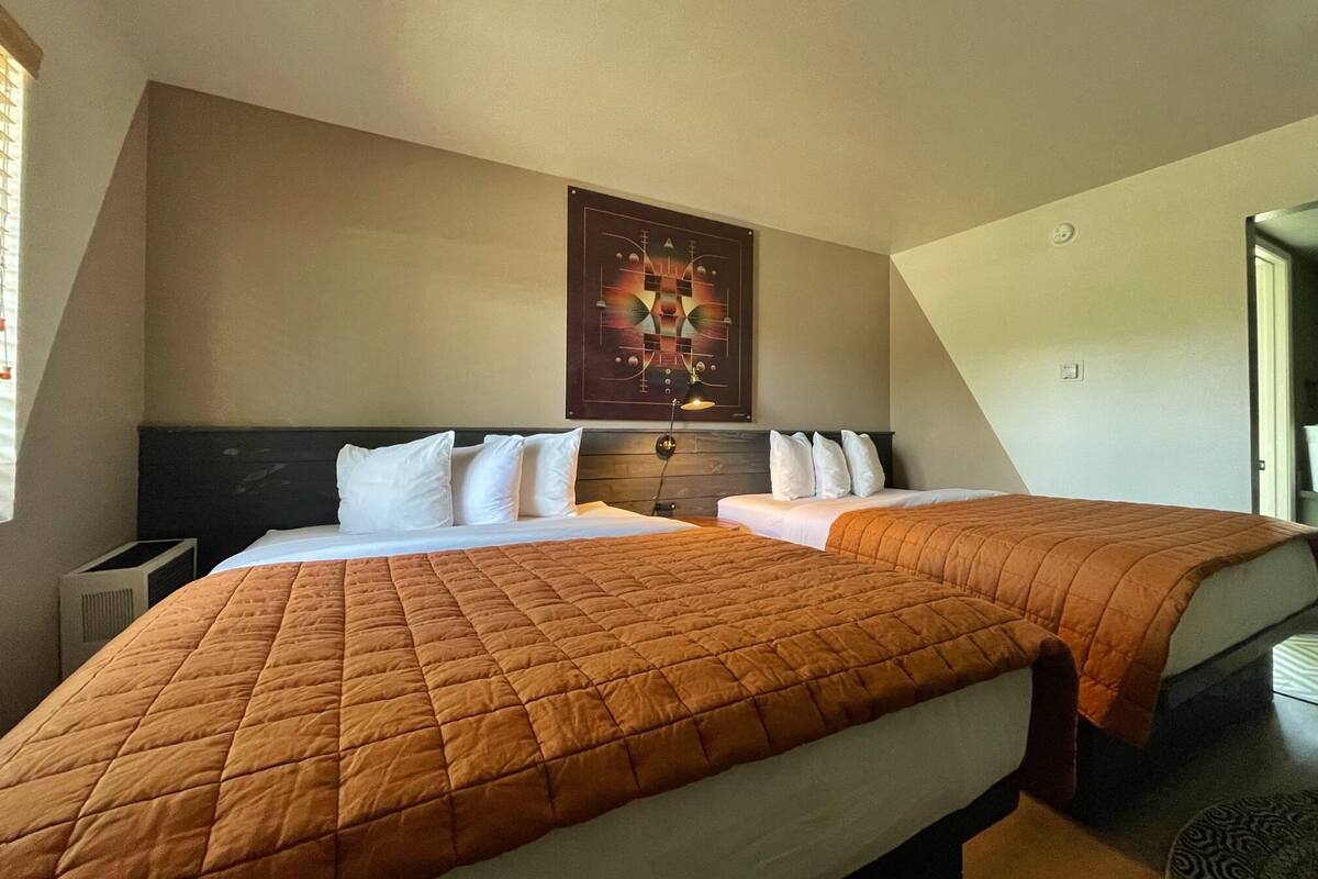 Quarto de hotel com duas camas, um quadro grande na parede e uma janela com persianas.