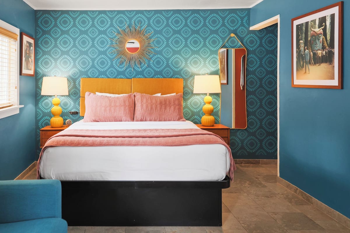 Quarto de hotel com cama, duas mesas de cabeceira com luminárias, um espelho grande e uma parede com desenho de raios de sol.
