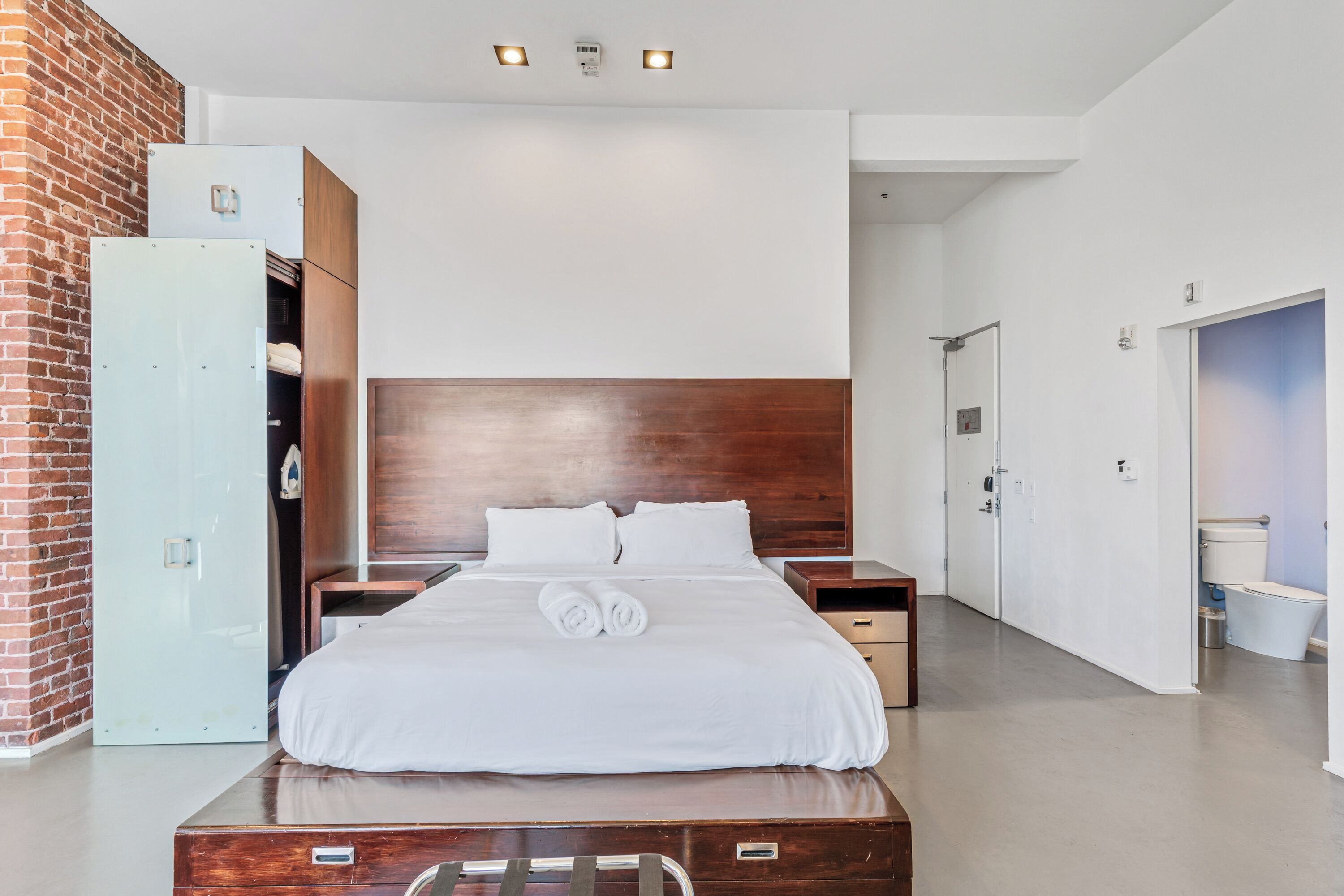 Quarto de hotel moderno com cabeceira de madeira, uma cama grande, um guarda-roupa e um banheiro visível por uma porta aberta.