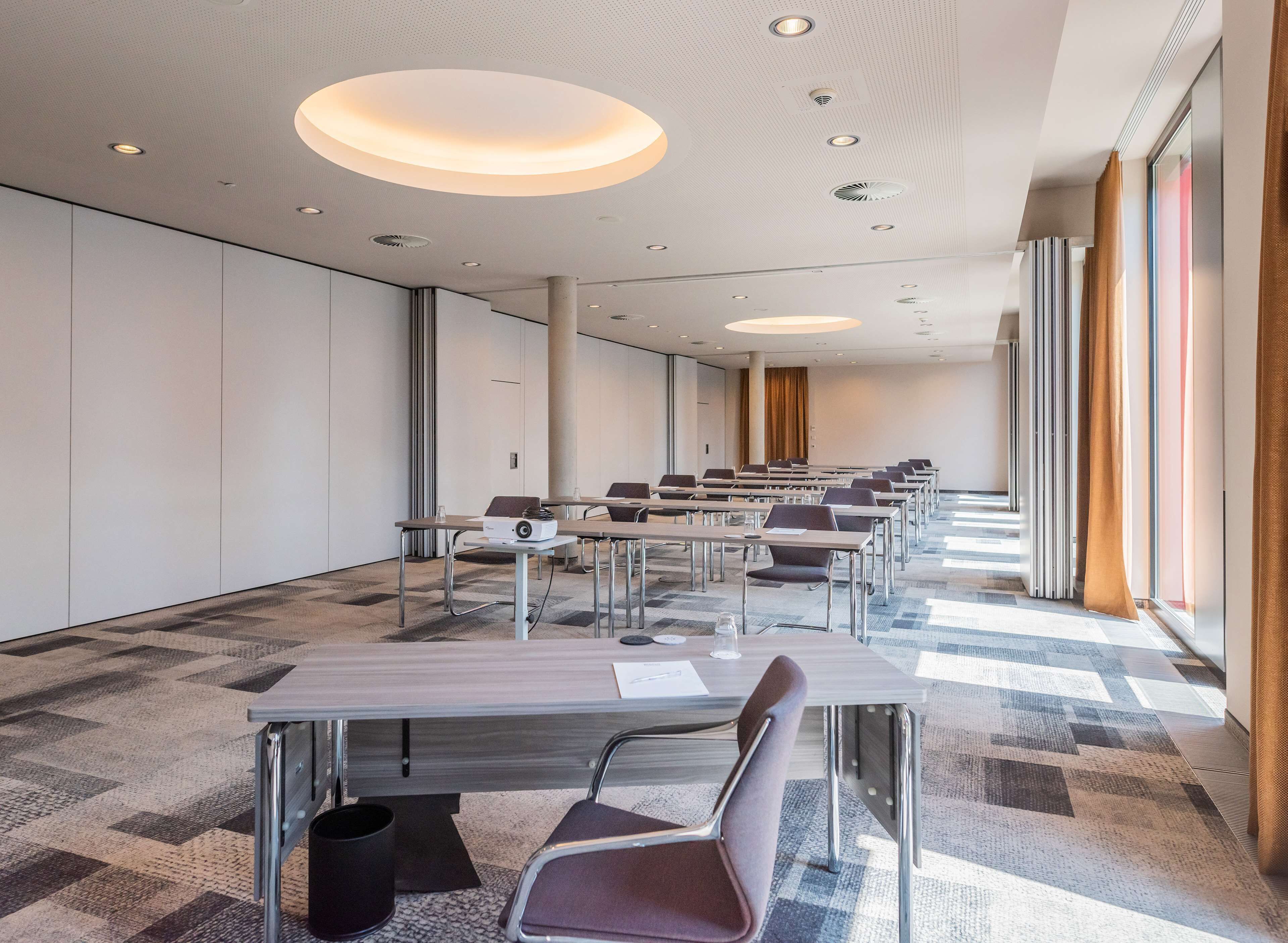 Uma sala de conferências com várias mesas e cadeiras, janelas amplas e um design de teto moderno.