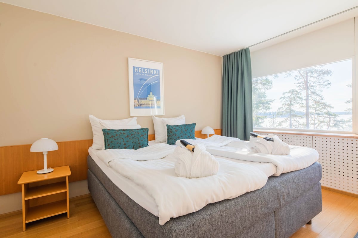 Quarto de hotel com uma cama grande, uma mesa de cabeceira, um cartaz de Helsínquia e uma janela com vista para árvores.