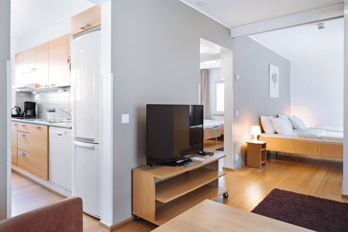 Apartamento moderno com cozinha, televisão em suporte de madeira, cama e pequena mesa de cabeceira com abajur.