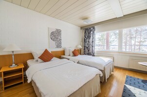 Quarto de hotel com duas camas, mesa de cabeceira, luminária, janela com cortinas e radiador.