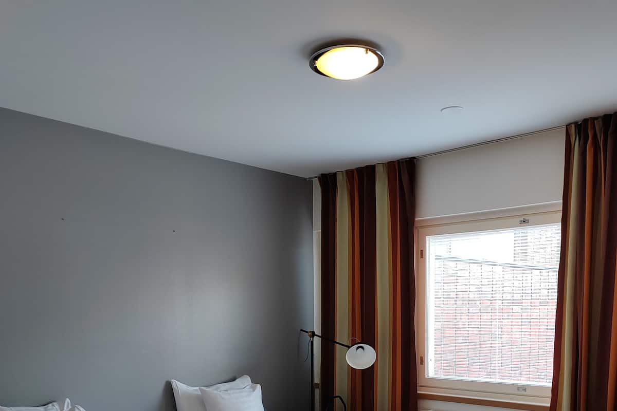 Quarto de hotel com duas camas de solteiro, janela com cortinas, luminária no teto e vista para prédios.