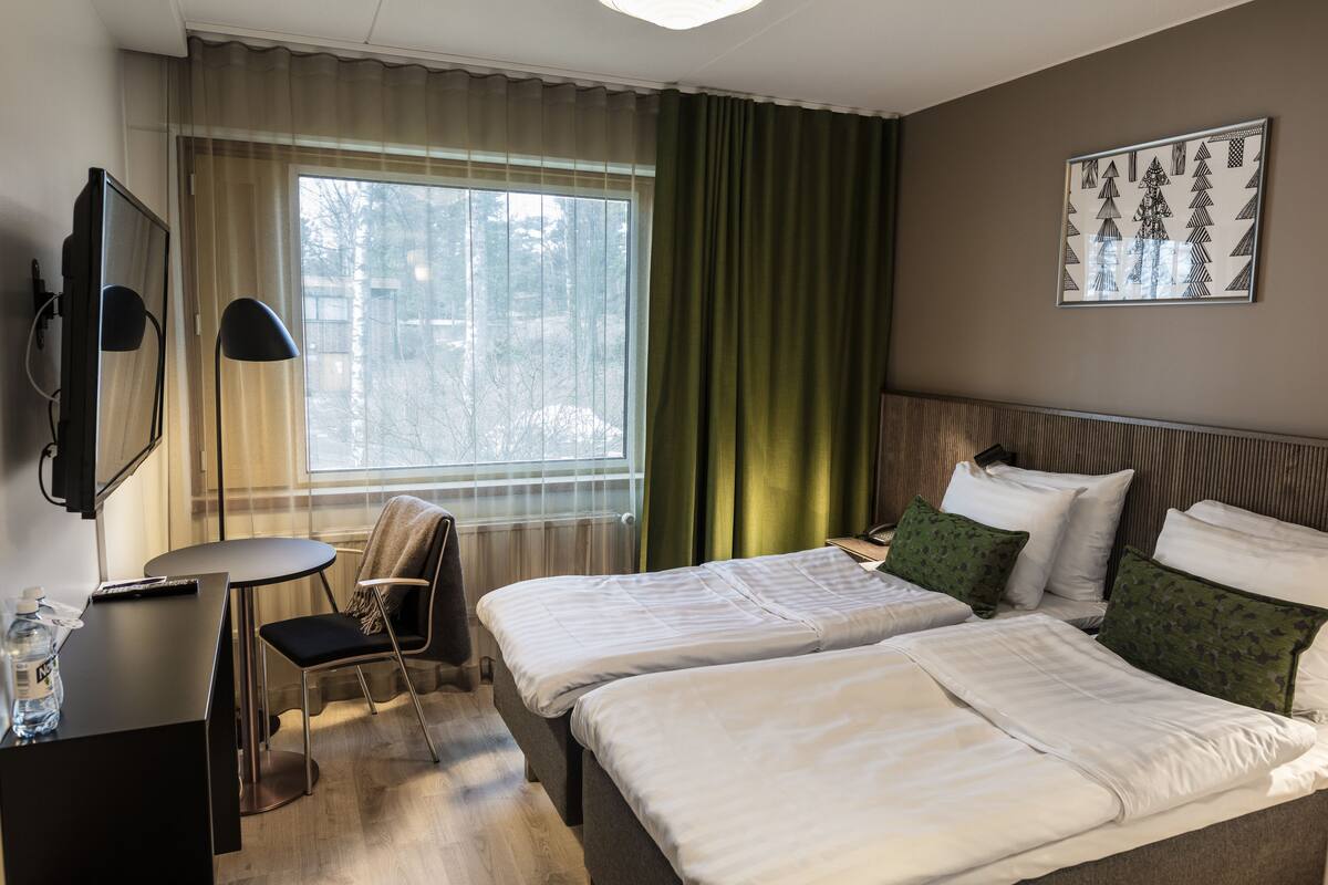 Quarto de hotel com duas camas, uma escrivaninha com cadeira, televisão e janela com cortinas.