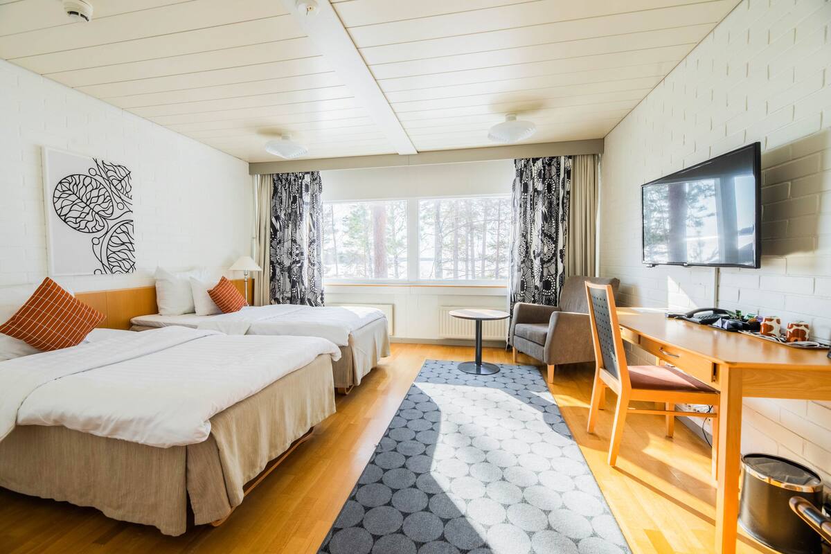 Quarto de hotel com duas camas, uma escrivaninha, uma cadeira, uma televisão e uma janela com cortinas.