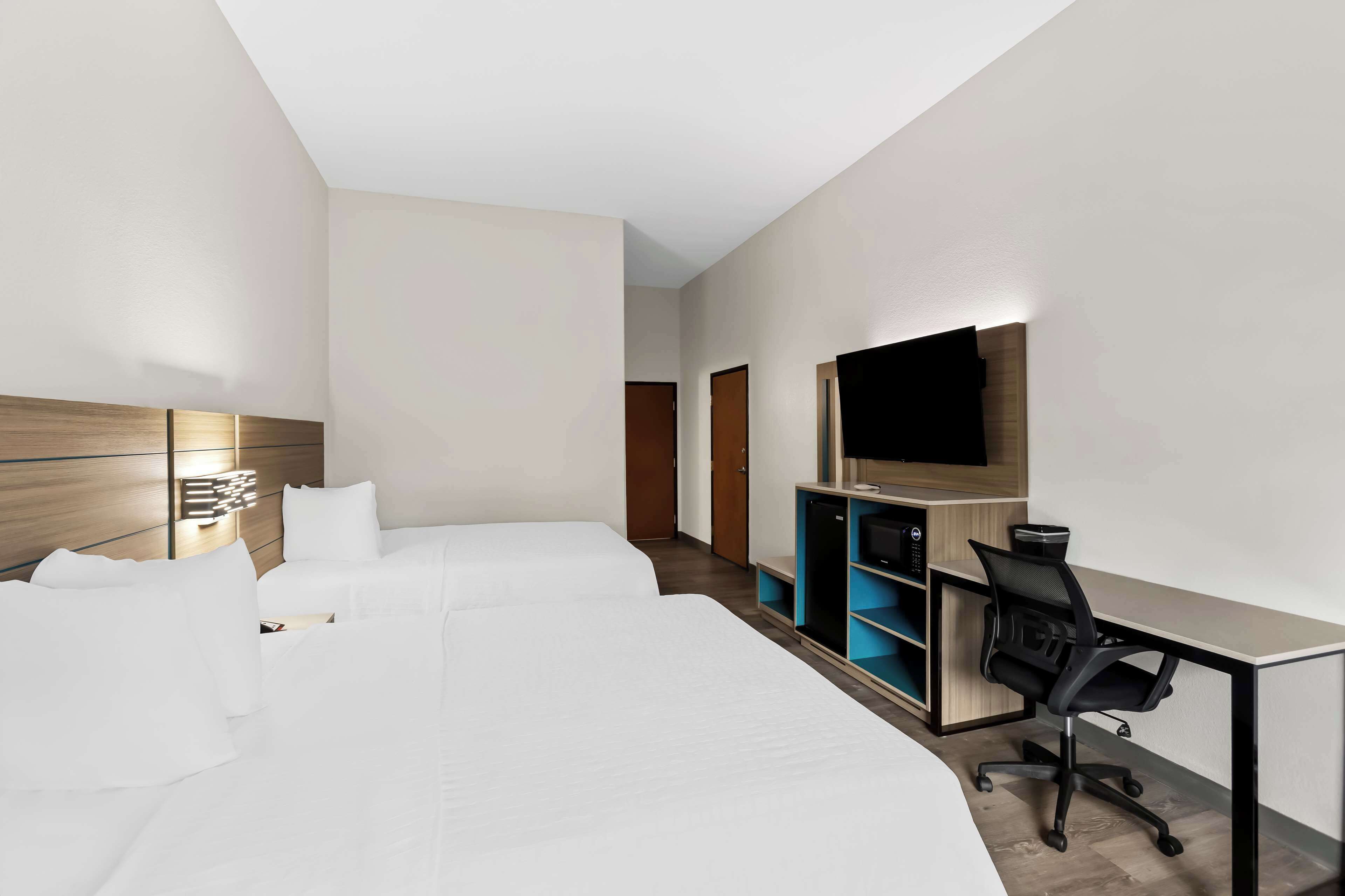 Quarto de hotel com duas camas, uma escrivaninha com cadeira, uma televisão e uma estante de livros.