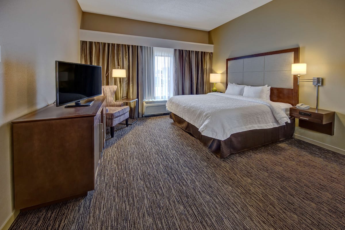 Quarto de hotel com uma cama grande, uma escrivaninha com televisão de tela plana, uma cadeira, uma mesinha de cabeceira com abajur e uma janela com cortinas.