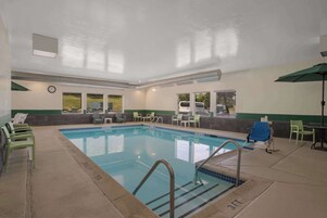 Piscina interior com água cristalina, rodeada por cadeiras verdes e brancas, um relógio na parede e um guarda-sol verde.