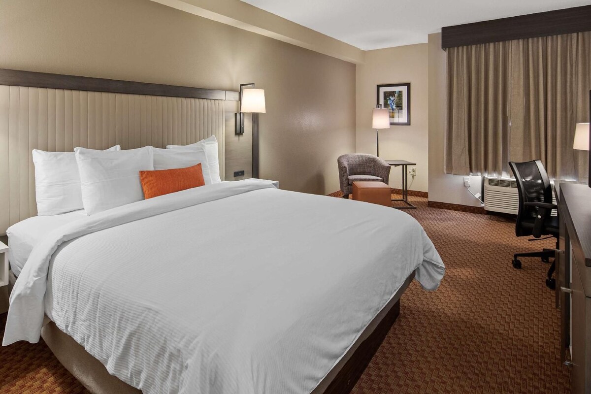 Quarto de hotel com uma cama grande, uma mesa com cadeira, uma televisão e uma pequena mesa com outra cadeira.