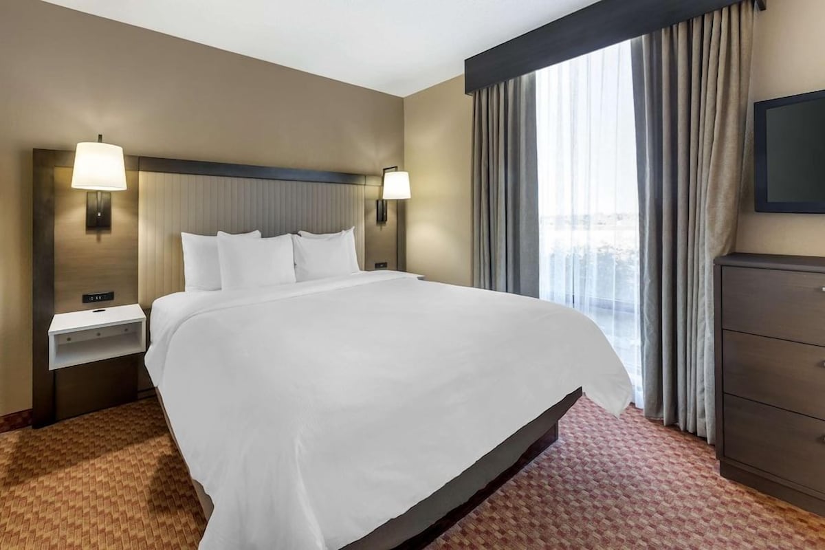 Quarto de hotel com uma cama grande, duas luminárias de cabeceira, mesa de cabeceira, cômoda e uma janela ampla com cortinas.
