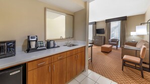 Quarto de hotel com cozinha compacta, área de jantar, sala de estar com sofá e poltrona, e televisão.
