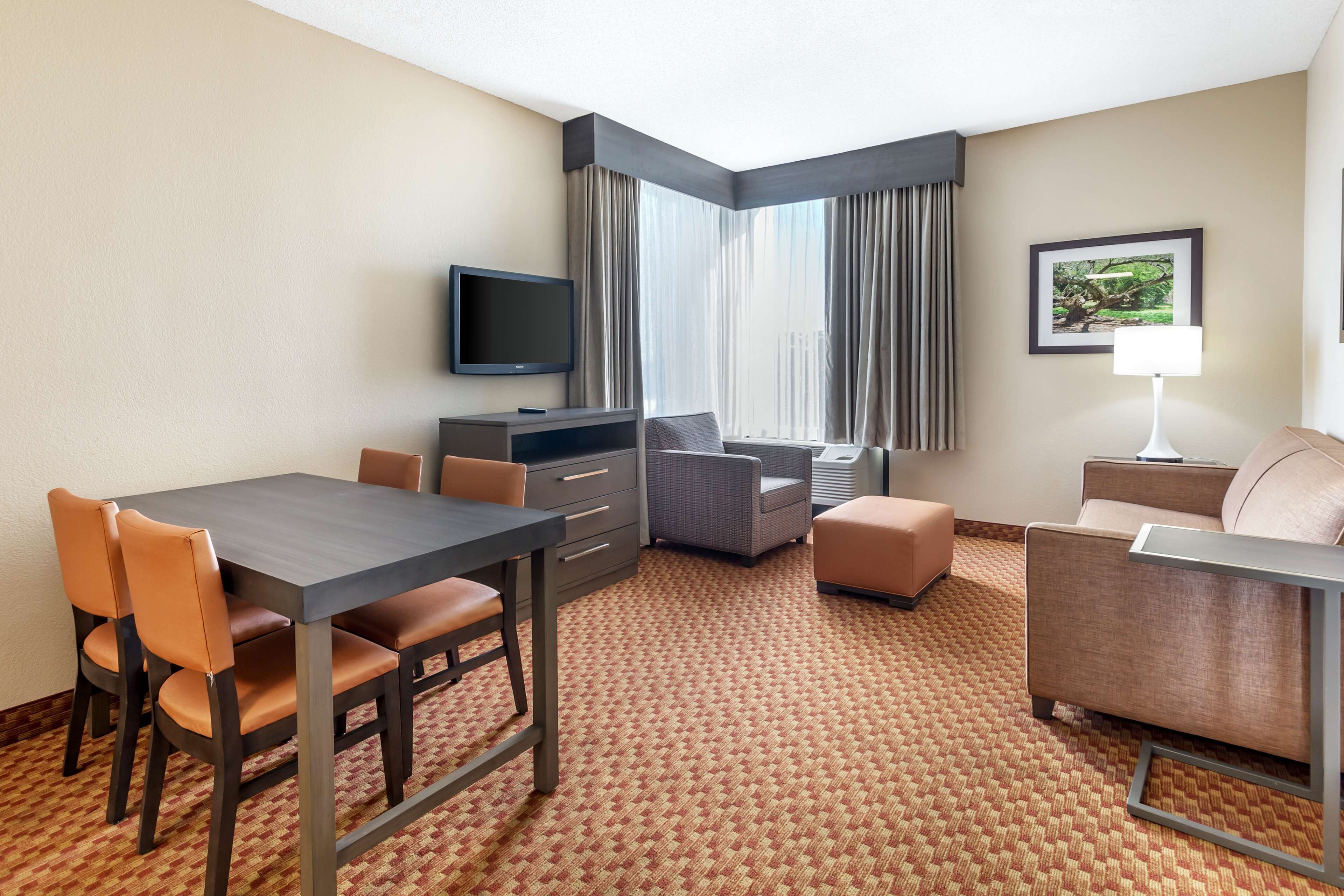 Quarto de hotel com mesa de jantar, cadeiras, sofá, televisão, luminária e janela com cortinas.