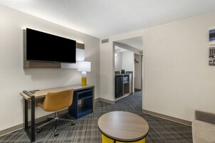 Quarto de hotel com televisão de tela plana na parede, uma escrivaninha com cadeira, uma mesinha e um micro-ondas ao fundo.