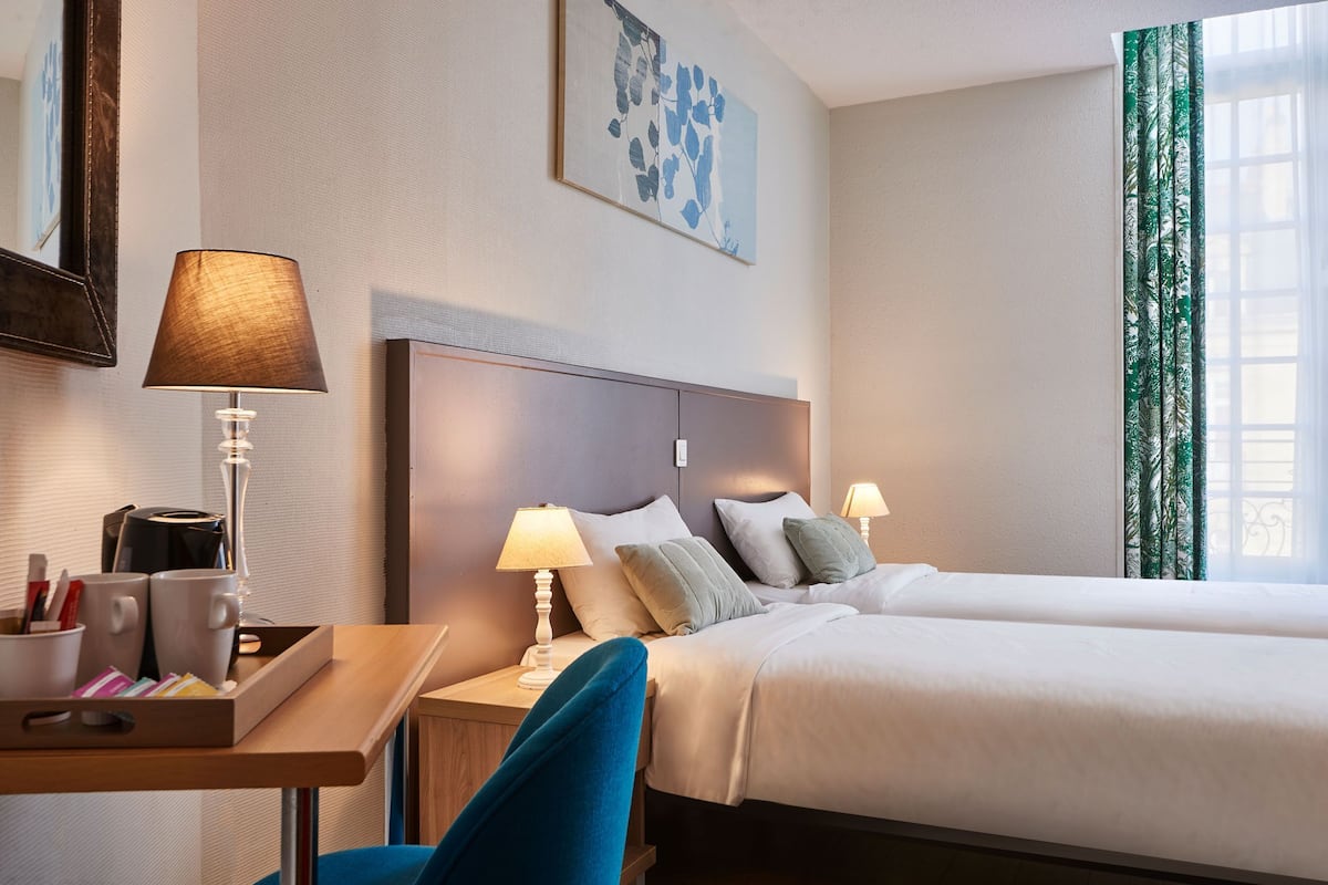 Quarto de hotel com uma cama, uma escrivaninha com luminária, um apoio de bandeja com xícaras e livros, e um quadro na parede.