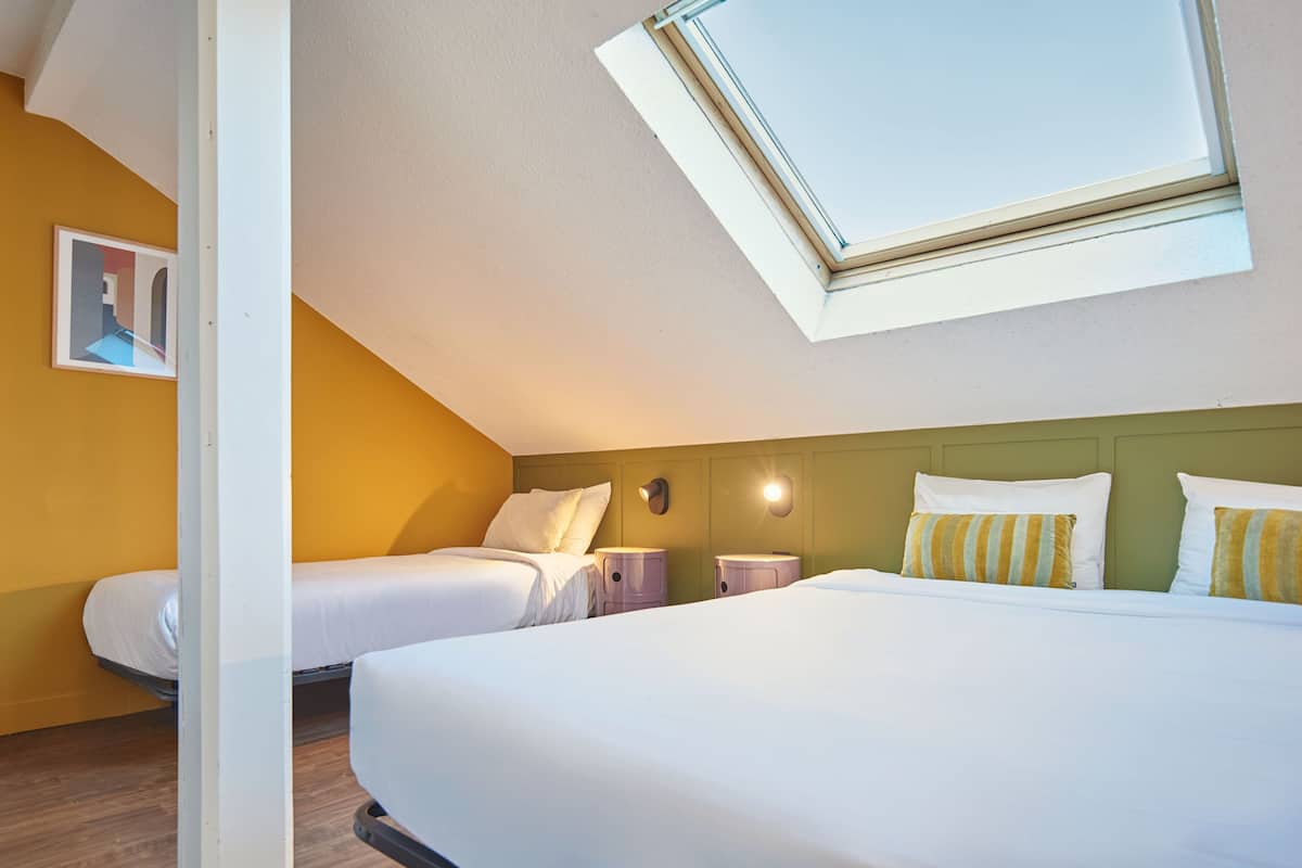 Quarto de hotel com duas camas, um claraboia, uma parede verde de destaque e um quadro na parede.