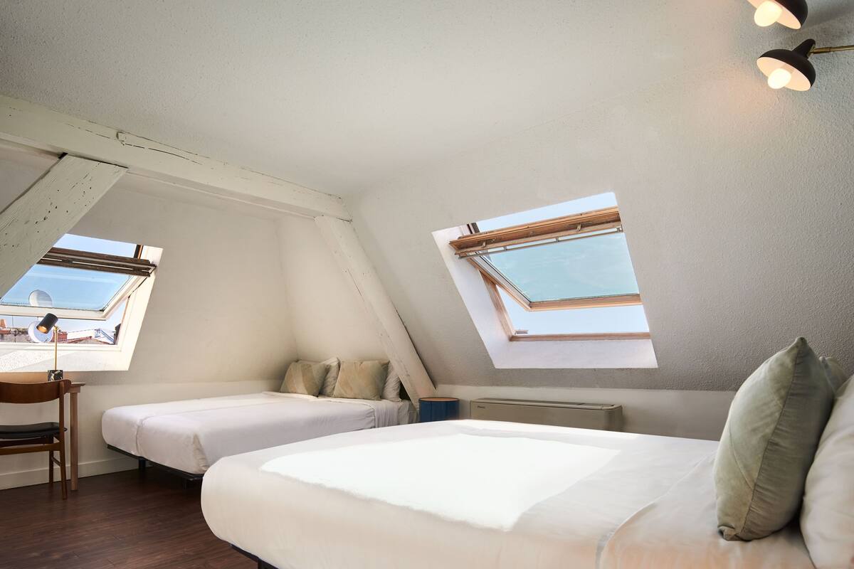 Quarto com duas camas, uma escrivaninha e claraboias.