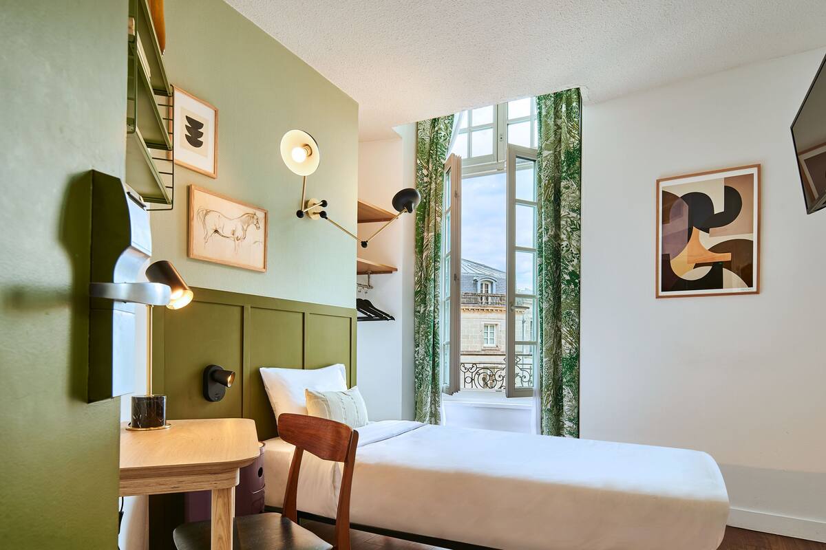 Quarto de hotel com cama, escrivaninha, cadeira, janela com cortinas e obras de arte nas paredes.