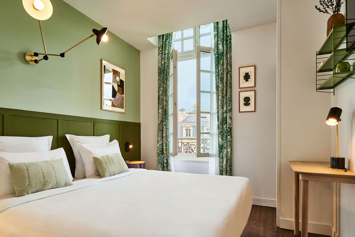 Quarto de hotel com uma cama grande, janela com cortinas verdes, mesa de cabeceira com abajur e prateleira na parede com objetos decorativos.