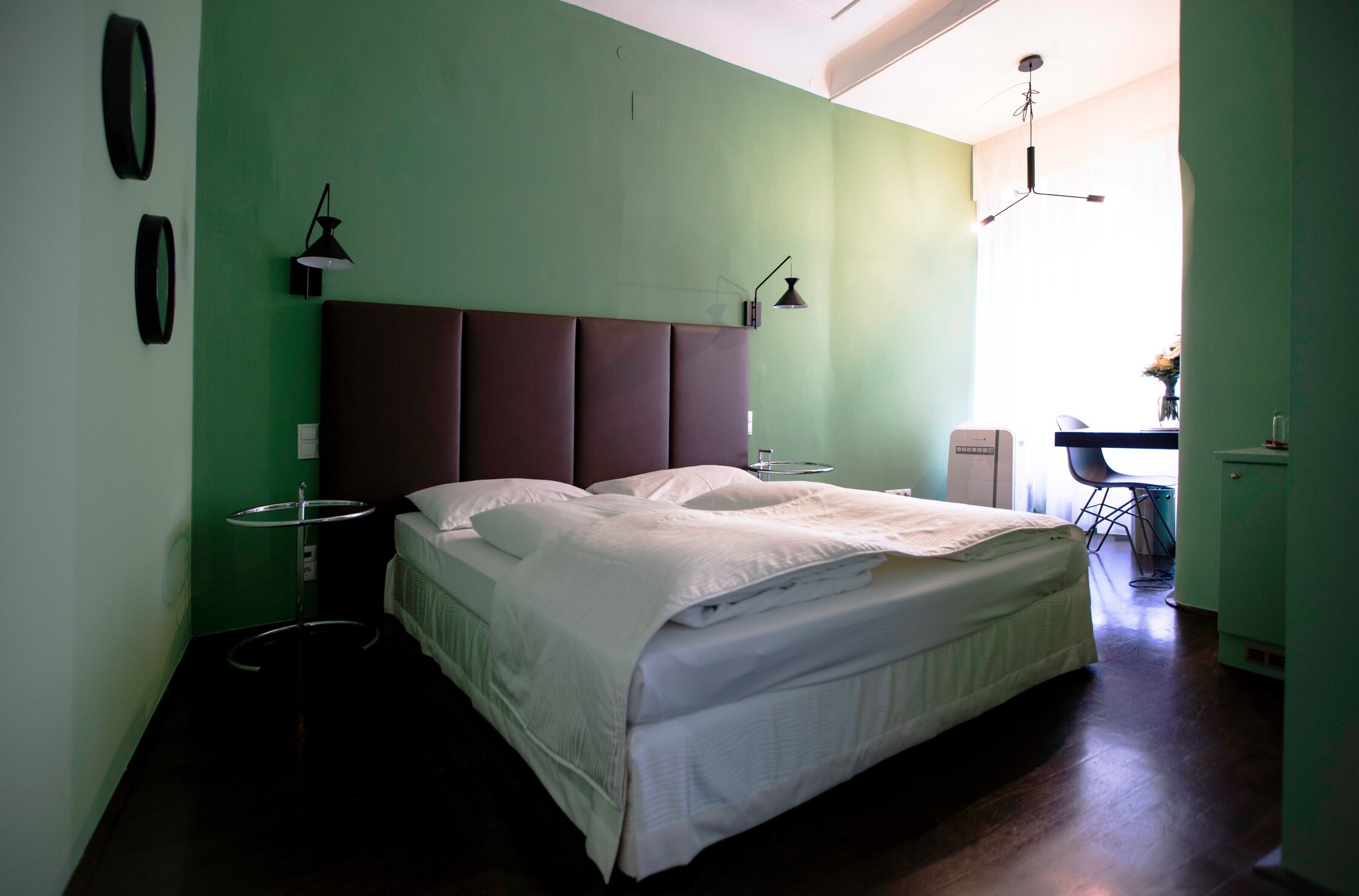 Cama bem arrumada em quarto com paredes verdes, soalho de madeira escura e uma secretária com cadeira ao fundo.